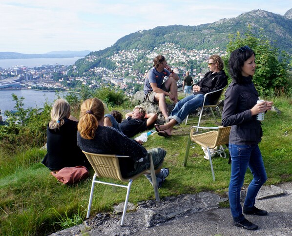 Um grupo de pessoas sentadas em cadeiras numa encosta gramada, com vista para uma cidade e um corpo de água.
