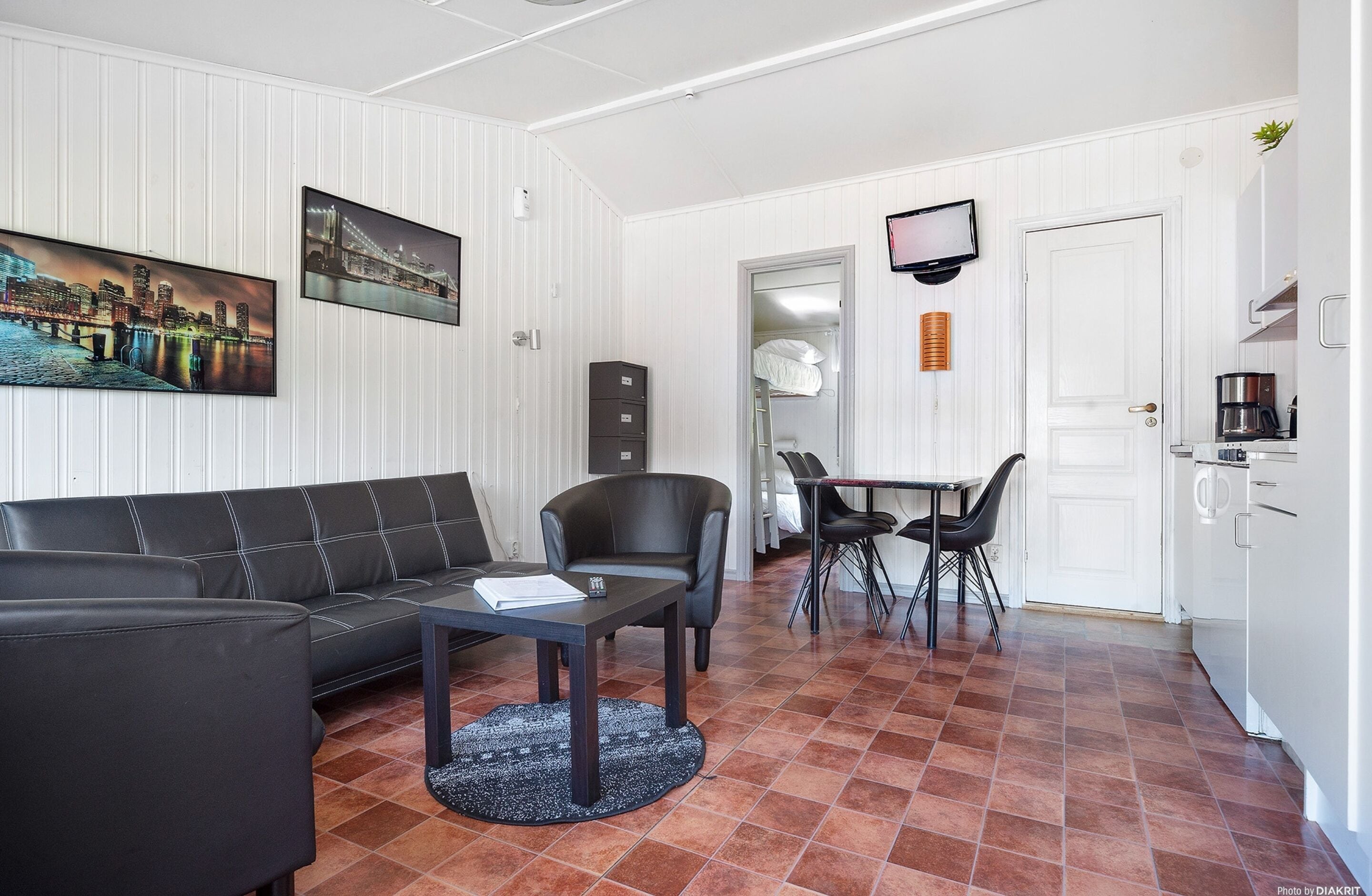 Uma sala de estar com sofá de couro preto, mesa de centro e TV fixada na parede. Há uma área de jantar com mesa e cadeiras, e uma cozinha com armários e eletrodomésticos.