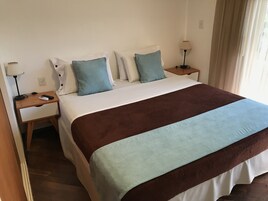 Cama bem arrumada com colcha marrom e azul clara, dois travesseiros iguais e uma mesa de cabeceira com abajur e controle remoto.