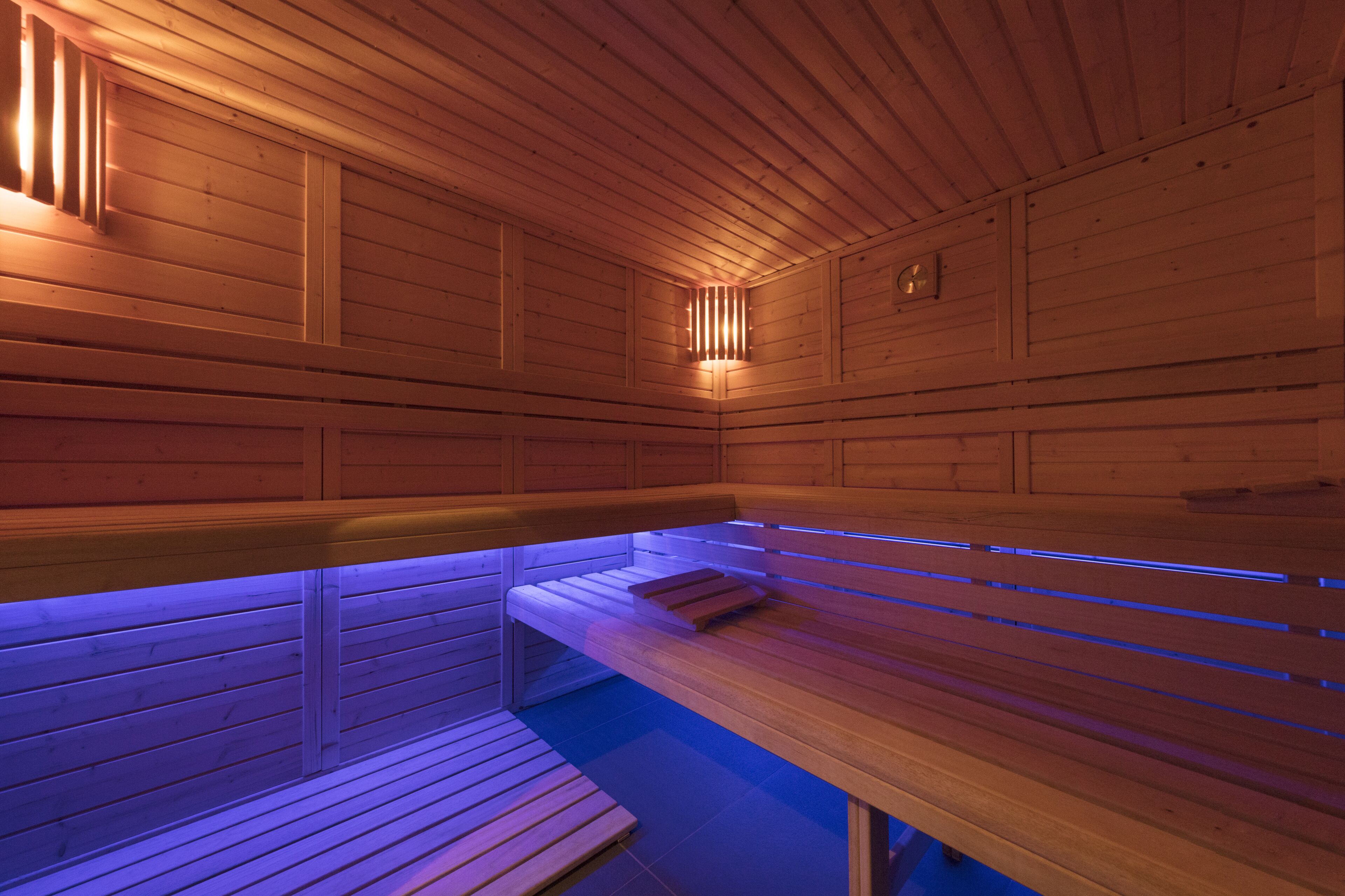 Uma sauna com bancos de madeira e piso inclinado, iluminada por luzes azuis e laranja.
