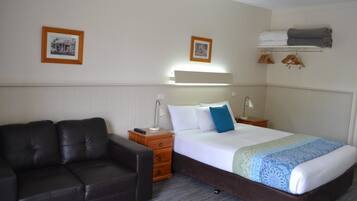 Quarto de hotel com cama, sofá de couro, mesa de cabeceira, abajur e prateleira na parede com toalhas.