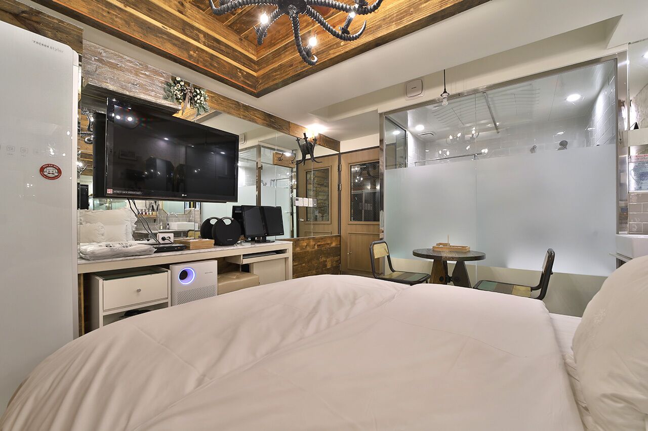 Quarto de hotel com cama, escrivaninha com computador, televisão e área de chuveiro.