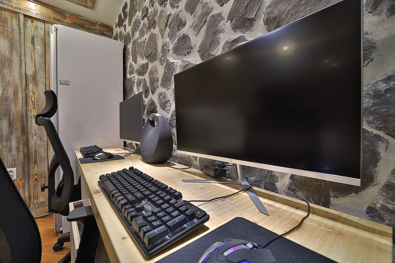 Um espaço de escritório em casa com monitor grande, teclado e rato sobre uma secretária de madeira, com parede de pedra ao fundo.