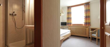 Quarto de hotel com chuveiro, cama, escrivaninha e janela com cortinas.