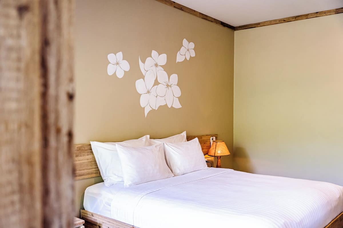 Quarto com cama, almofadas brancas, cabeceira de madeira, parede com adesivos florais e abajur.