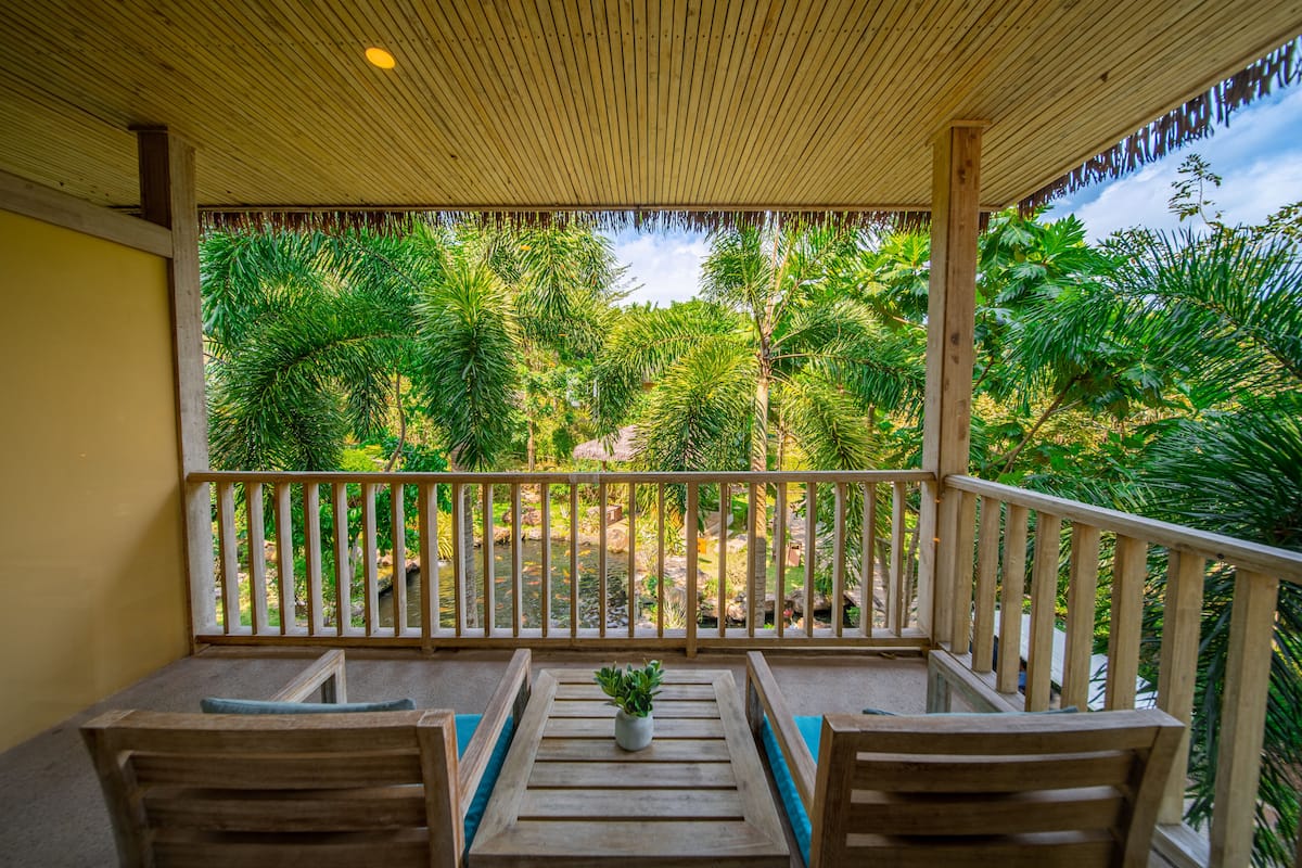 Um deck de madeira com guarda-corpo, duas cadeiras de madeira, uma pequena mesa com uma planta e vista para vegetação exuberante e um lago.