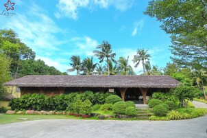Um edifício com telhado de palha e um jardim na frente, rodeado por palmeiras e outras plantas tropicais.