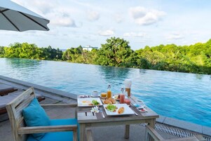 Mesa de jantar perto de uma piscina, com vista para vegetação exuberante e um edifício ao fundo.