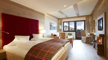 Quarto de hotel com uma cama grande, cabeceira vermelha, área de estar com poltrona e mesa, lareira e vista para o exterior.