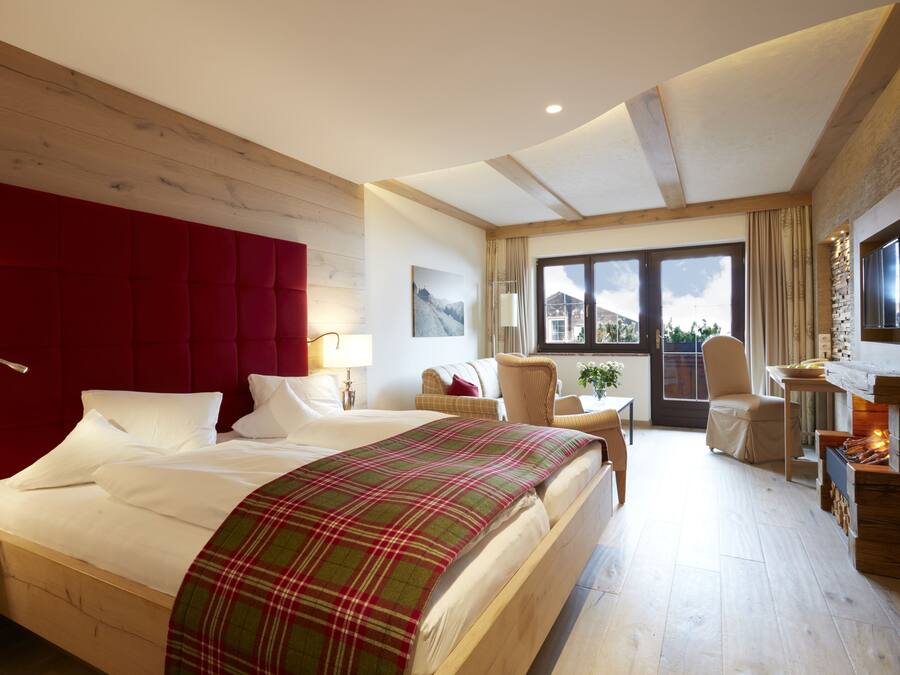Quarto de hotel com uma cama grande, cabeceira vermelha, área de estar com poltrona e mesa, lareira e vista para o exterior.