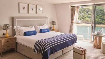 Quarto de hotel com uma cama grande, duas mesas de cabeceira com luminárias, vista para o mar e uma varanda com mesa e cadeiras.