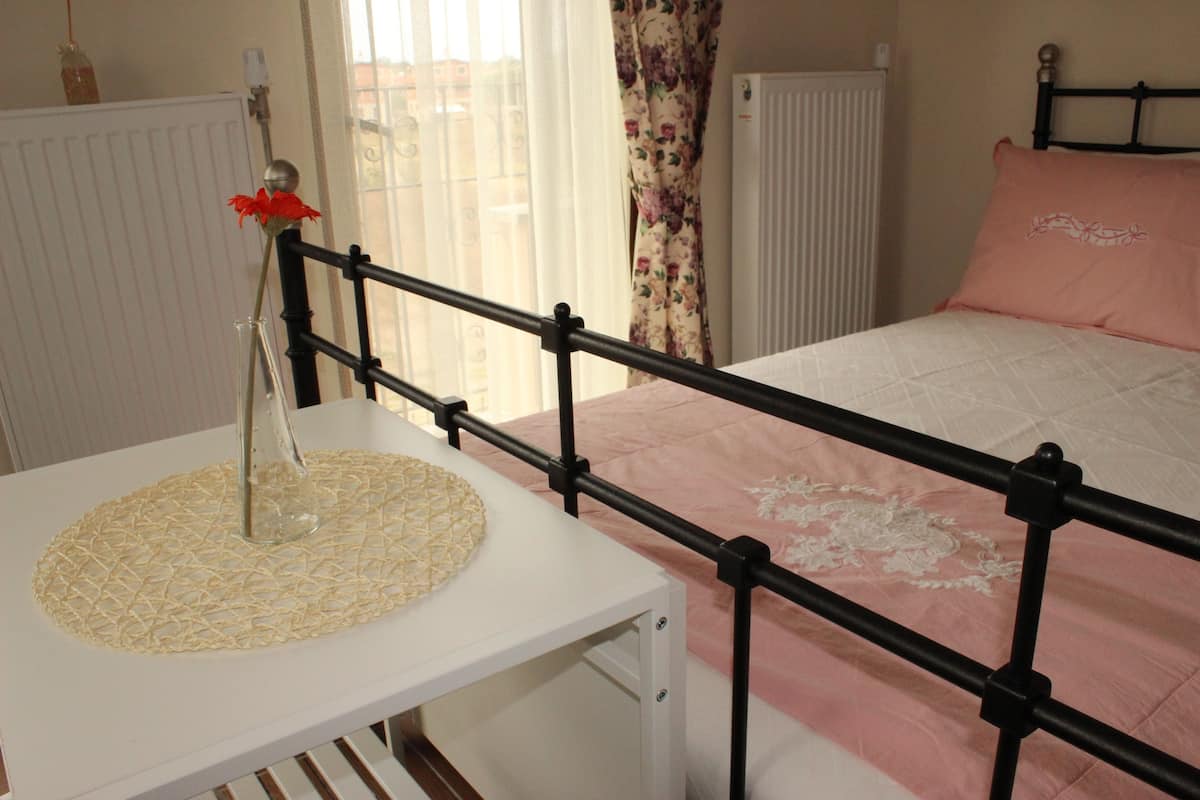 Quarto com cama, mesa de cabeceira, radiador e janela com cortinas.