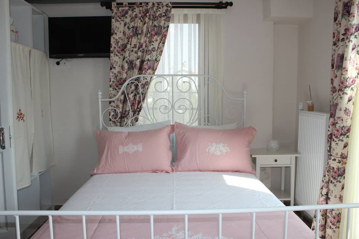Quarto com estrutura de cama de metal branca, duas almofadas cor-de-rosa, mesa de cabeceira branca e TV de tela plana na parede.