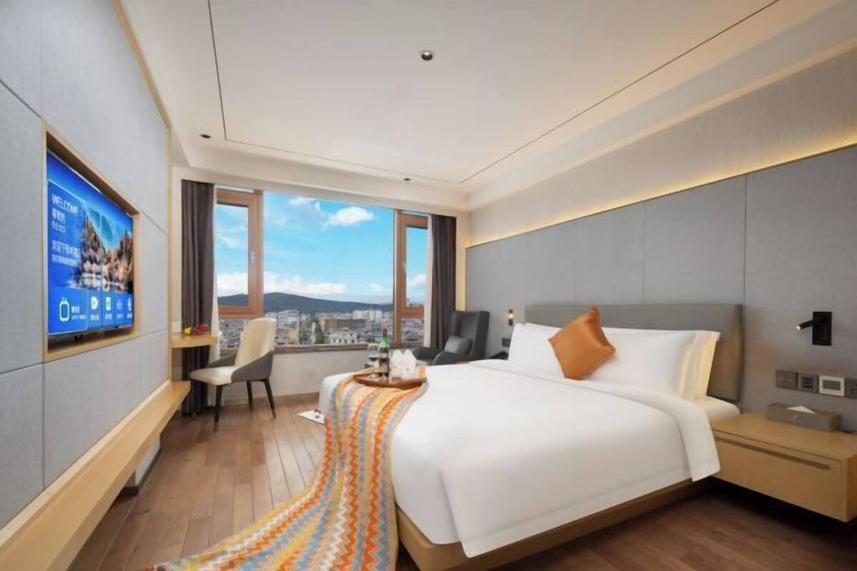 Quarto de hotel com cama, escrivaninha, poltrona, televisão e vista para a cidade.