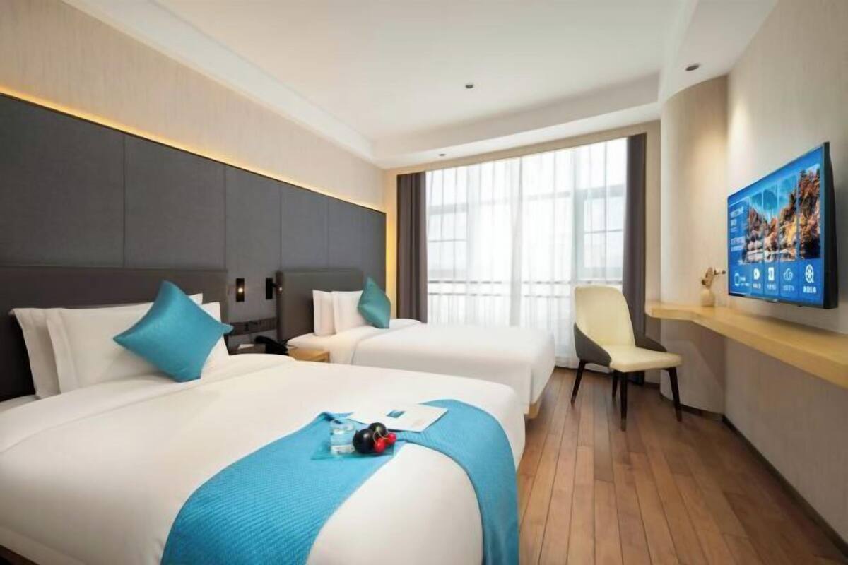 Quarto de hotel com duas camas, uma televisão, uma cadeira e uma janela grande.