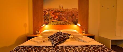 Quarto de hotel com uma cama grande, duas luminárias de cabeceira, um edredom estampado e uma pintura com vista para a cidade na parede.
