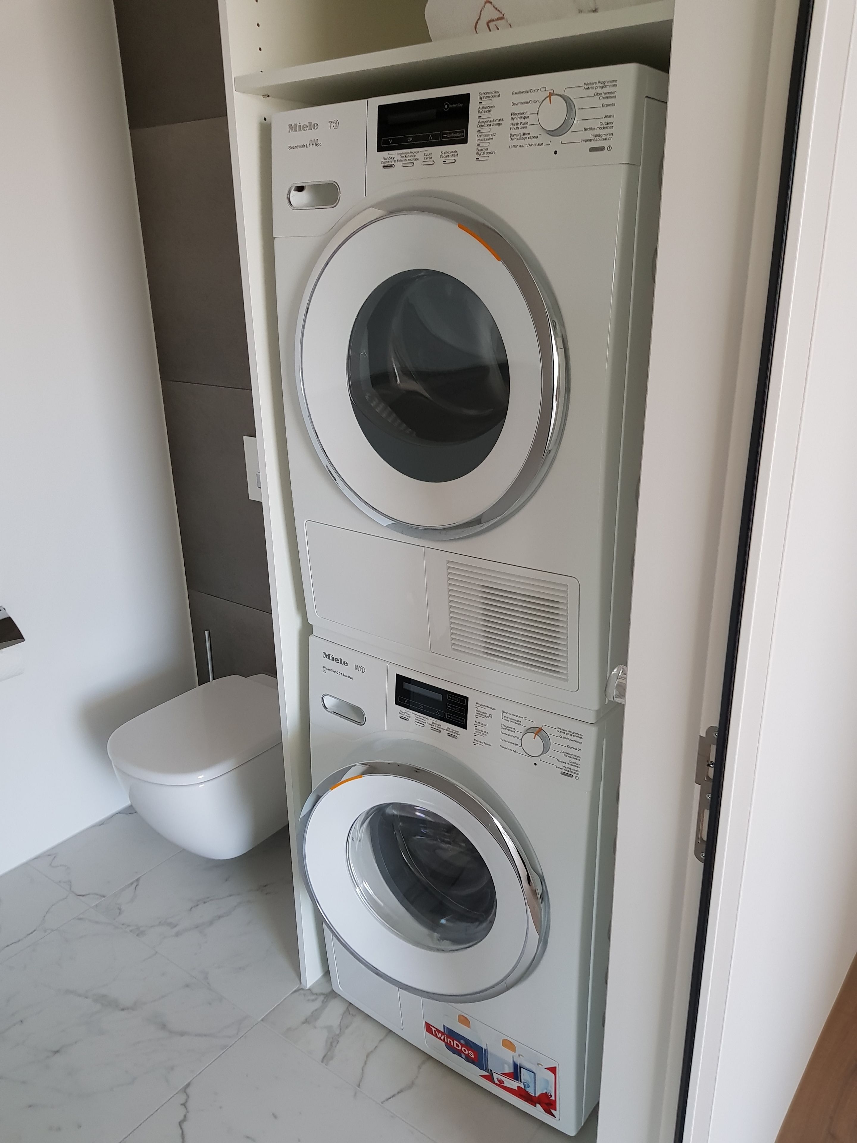 Uma máquina de lavar e secar compacta, fixada na parede, com duas portas, painel de controle digital e um pequeno compartimento de armazenamento.