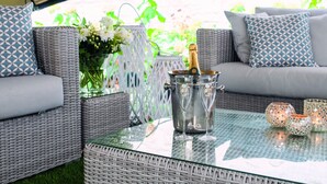 Um sofá de vime com almofadas estampadas, uma mesa de vidro com balde de champanhe e copos, e luzes decorativas.