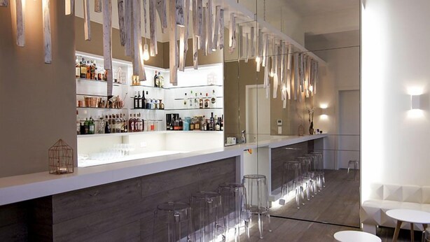 Uma área de bar moderna com balcão de madeira, prateleiras com garrafas e banquetas transparentes.