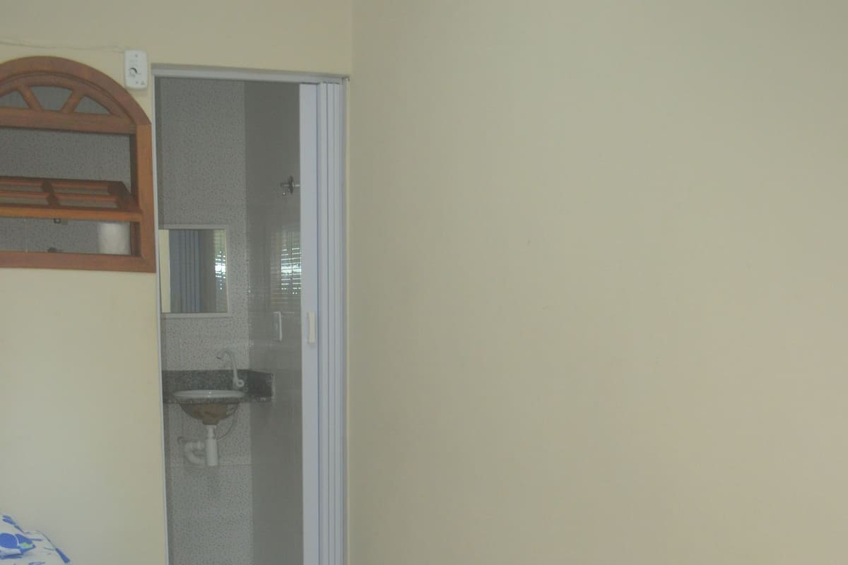 Quarto com duas camas, uma com colcha floral e outra com colcha azul florida. Um banheiro é visível através de uma porta de vidro.