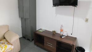 Quarto com televisão na parede, armário de madeira com gavetas, guarda-roupa cinzento e sofá bege.