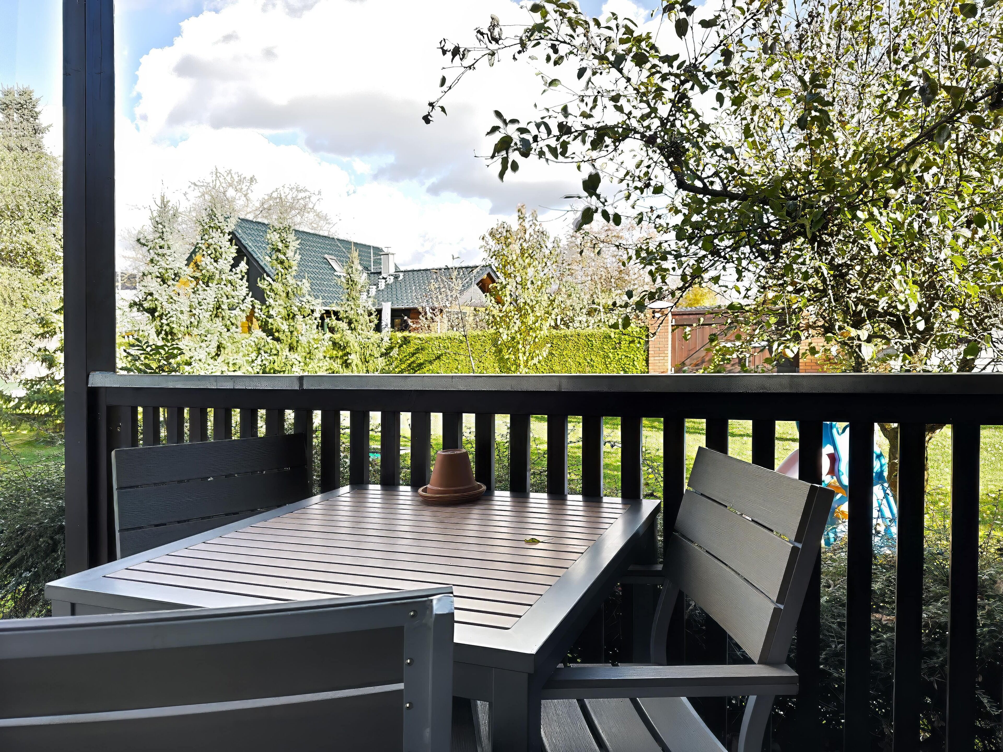 Um deck de madeira com uma mesa e cadeiras, um vaso de planta e vista para um jardim e edifícios.