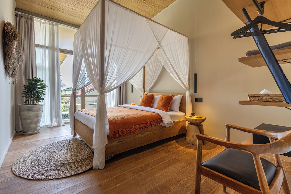 Quarto com cama de dossel, piso de madeira, tapete redondo tecido, cadeira de madeira e uma grande planta em vaso.