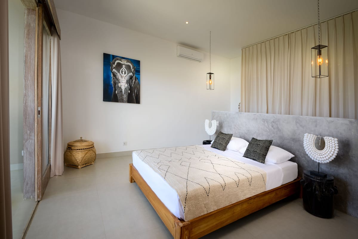 Quarto com uma cama grande, ar condicionado fixado na parede, luminária pendente e um detalhe decorativo na parede.