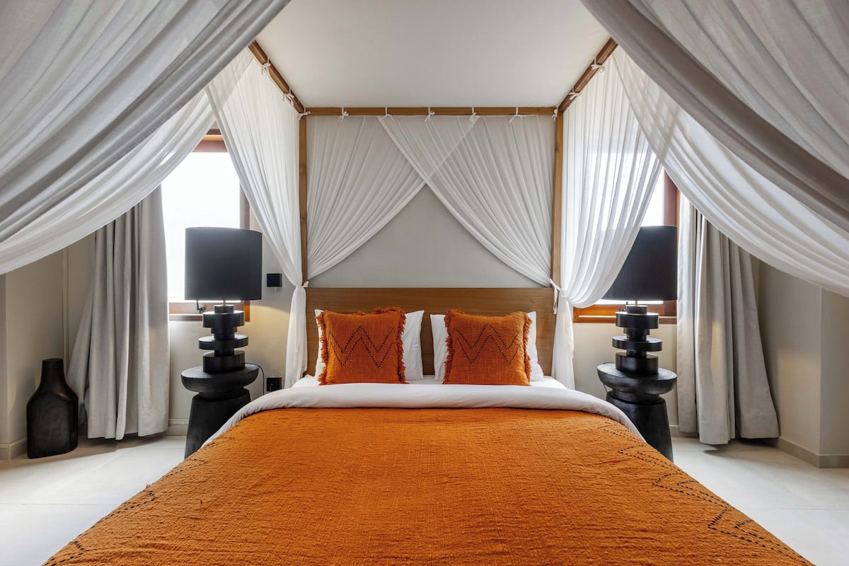 Uma cama com roupa de cama branca e laranja, duas mesas de cabeceira com abajures e cortinas leves.
