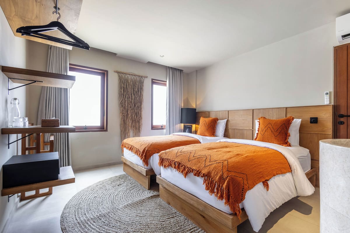 Quarto moderno com duas camas, cabeceira de madeira, mesa de cabeceira, luminária pendente e janela com cortinas.