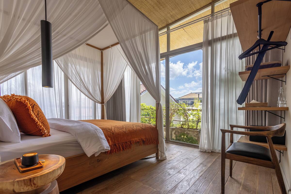 Quarto com cama, cadeira, mesa e uma grande janela com cortinas.