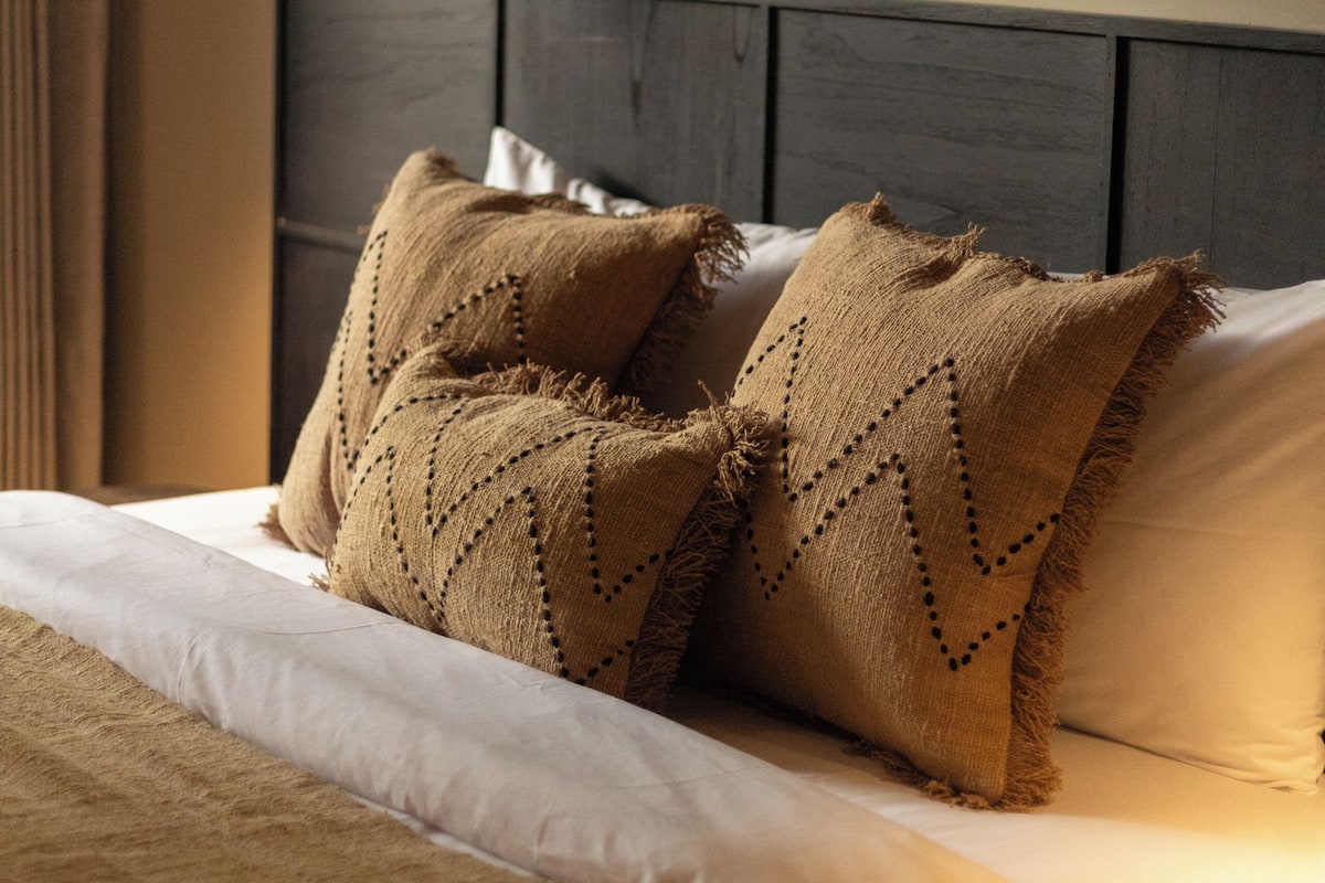 Uma cama com cabeceira escura, duas almofadas decorativas e um cobertor claro.