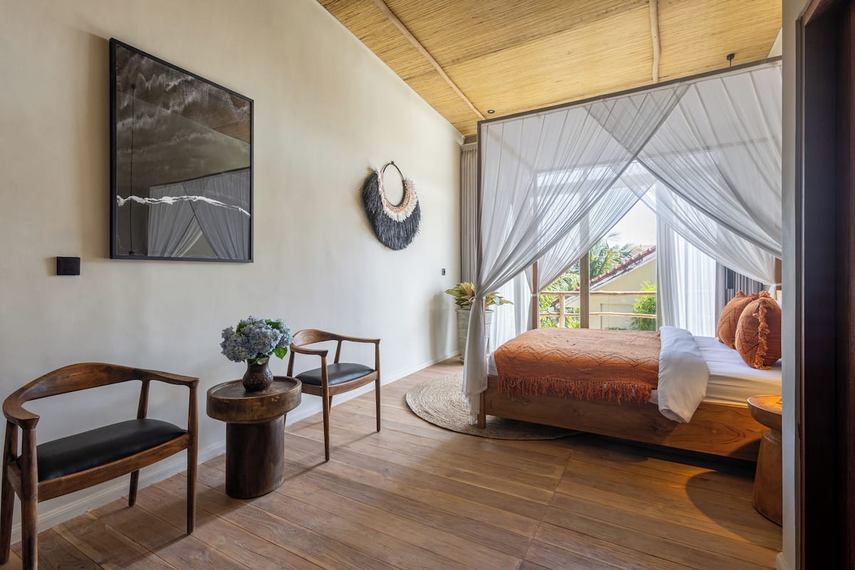 Quarto com cama, duas cadeiras, uma mesa redonda, um espelho grande e vista através de cortinas translúcidas.