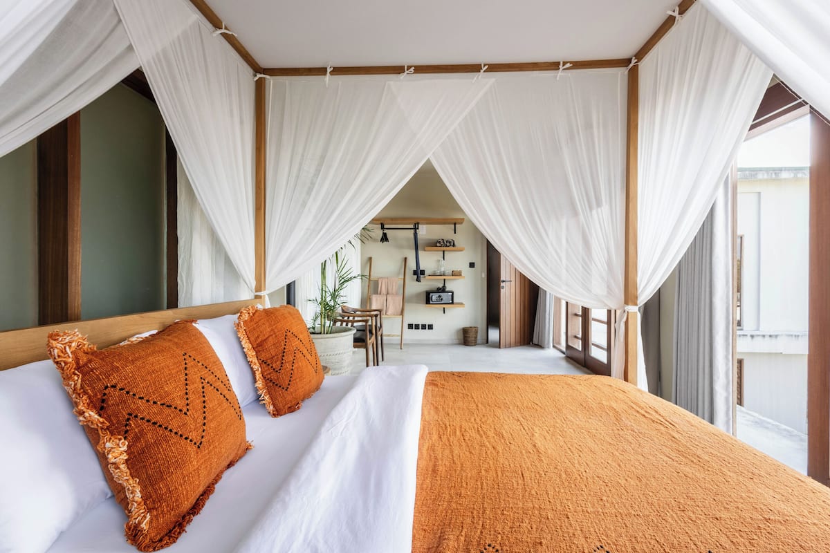 Quarto com cama, cortinas brancas e vista para outro espaço com planta e mesa.