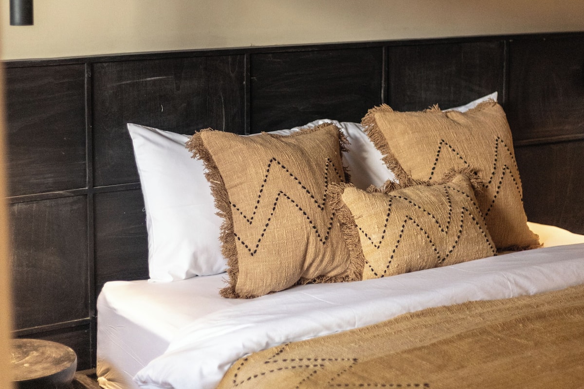 Cama com almofadas brancas e estampadas, cabeceira escura e duas molduras na parede.