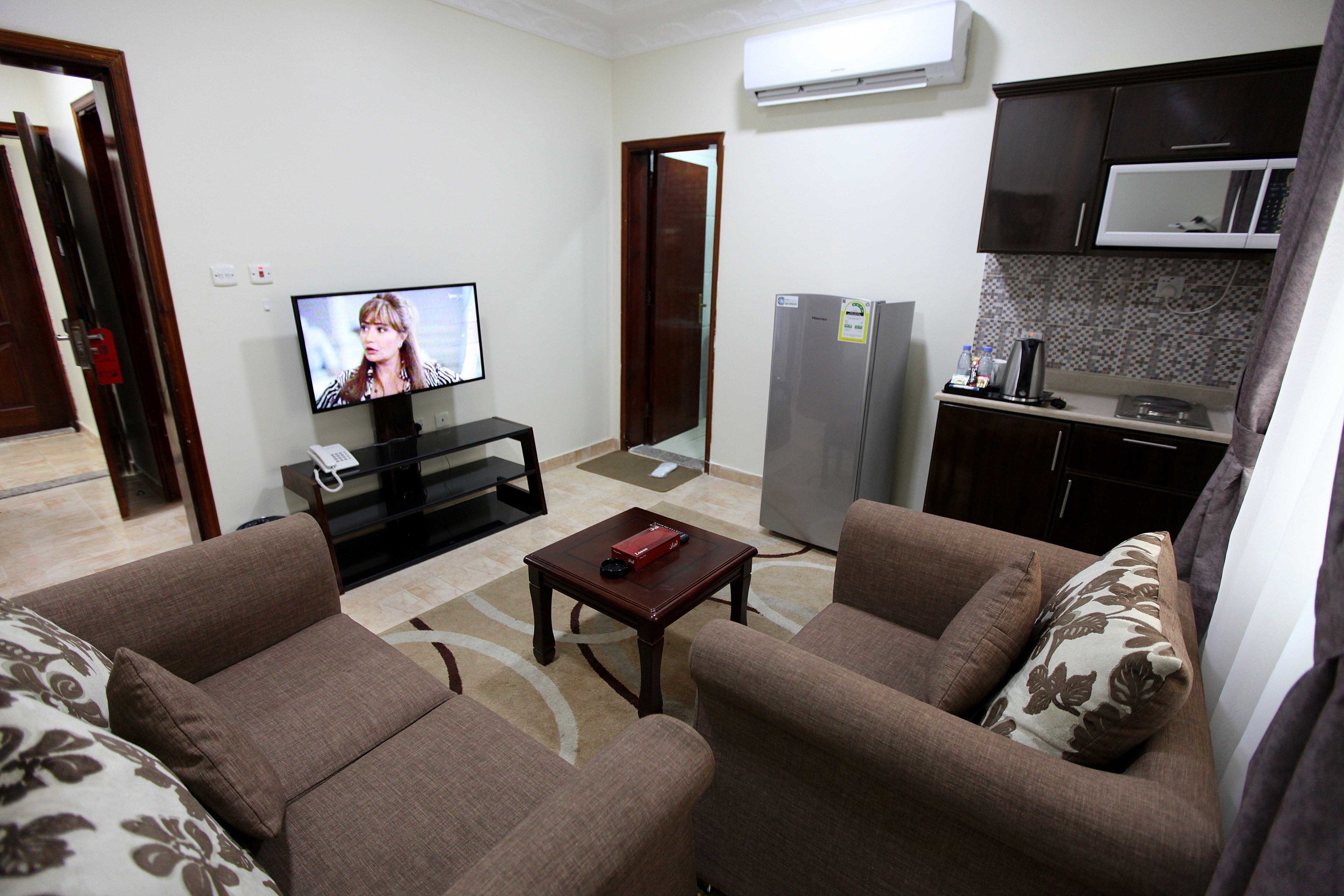 Uma sala de estar com sofá, mesa de centro, televisão, geladeira e cozinha equipada com armários e micro-ondas.