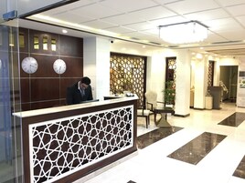 Uma área de recepção de hotel com recepcionista, balcão de atendimento, dois relógios na parede, divisória decorativa e um lustre moderno.