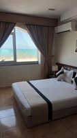 Quarto de hotel com uma cama grande, mesa de cabeceira, abajur e vista para a praia.