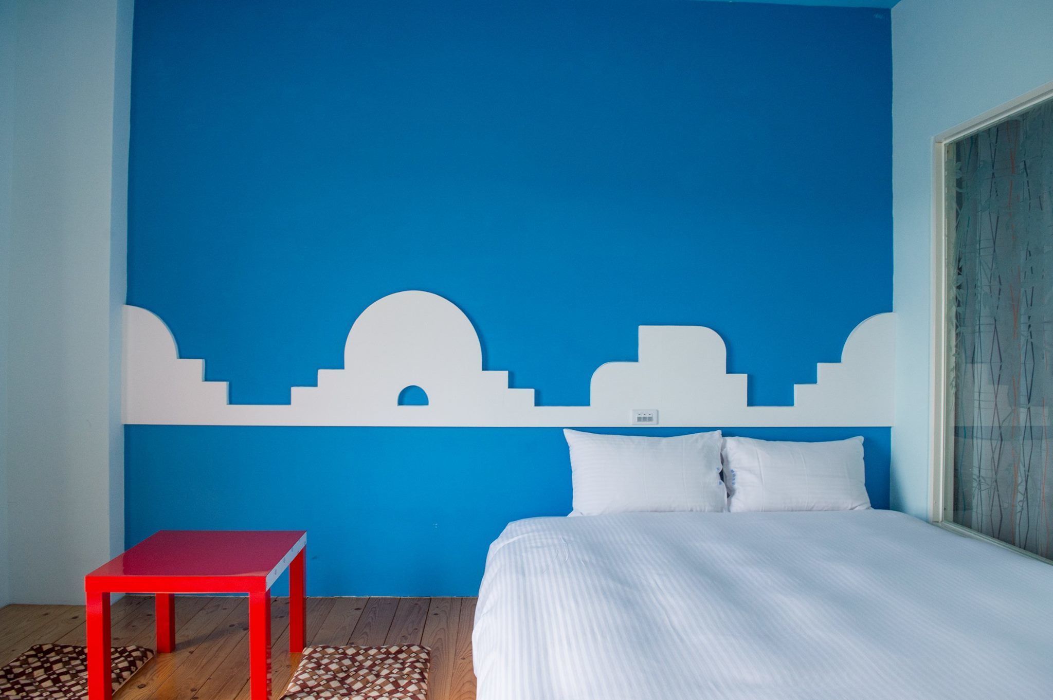 Quarto com uma cama, uma mesa vermelha e uma parede azul com padrões geométricos brancos.