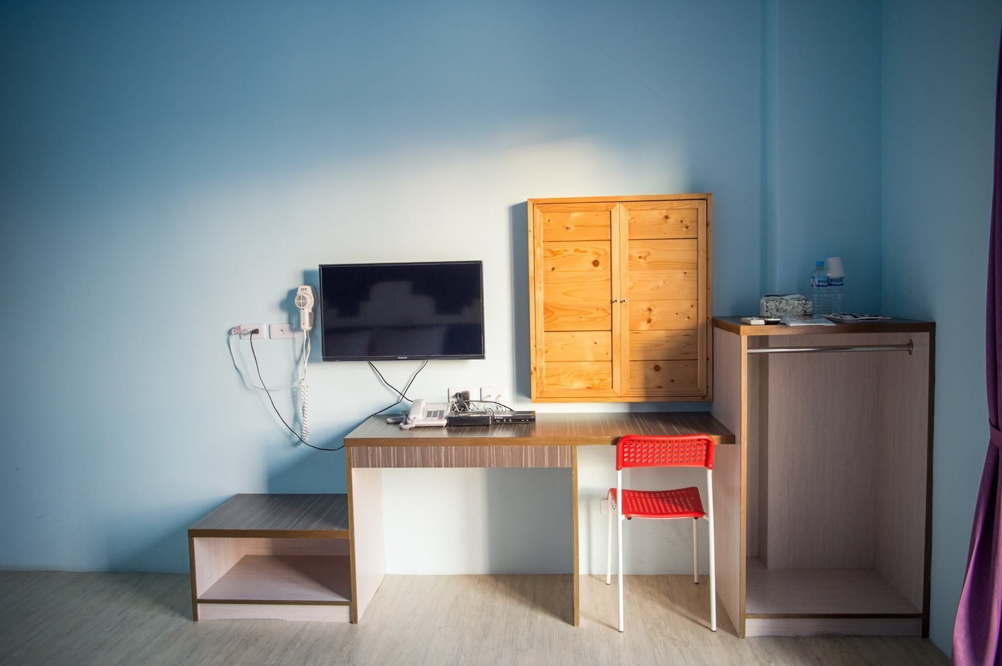 Quarto com escrivaninha de madeira, cadeira vermelha, televisão na parede, guarda-roupa de madeira e uma pequena mesa com uma garrafa de água.