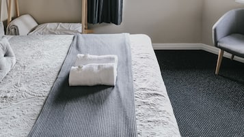 Quarto de hotel com cama, ventoinha no teto, janela com cortinas, uma cadeira e cabideiro.