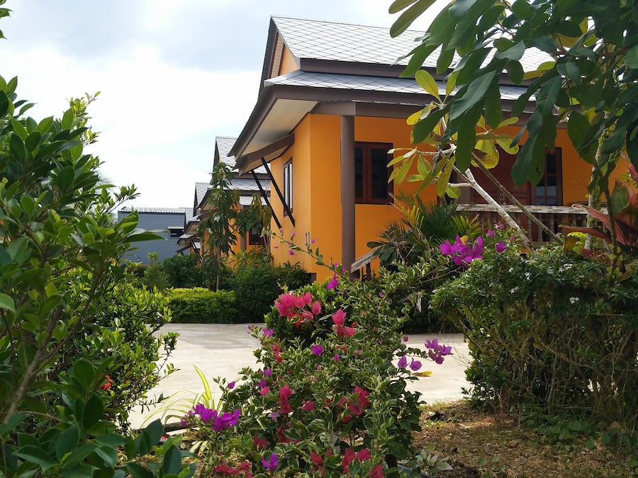 Um edifício amarelo com telhado inclinado, rodeado por vegetação exuberante e plantas floridas.