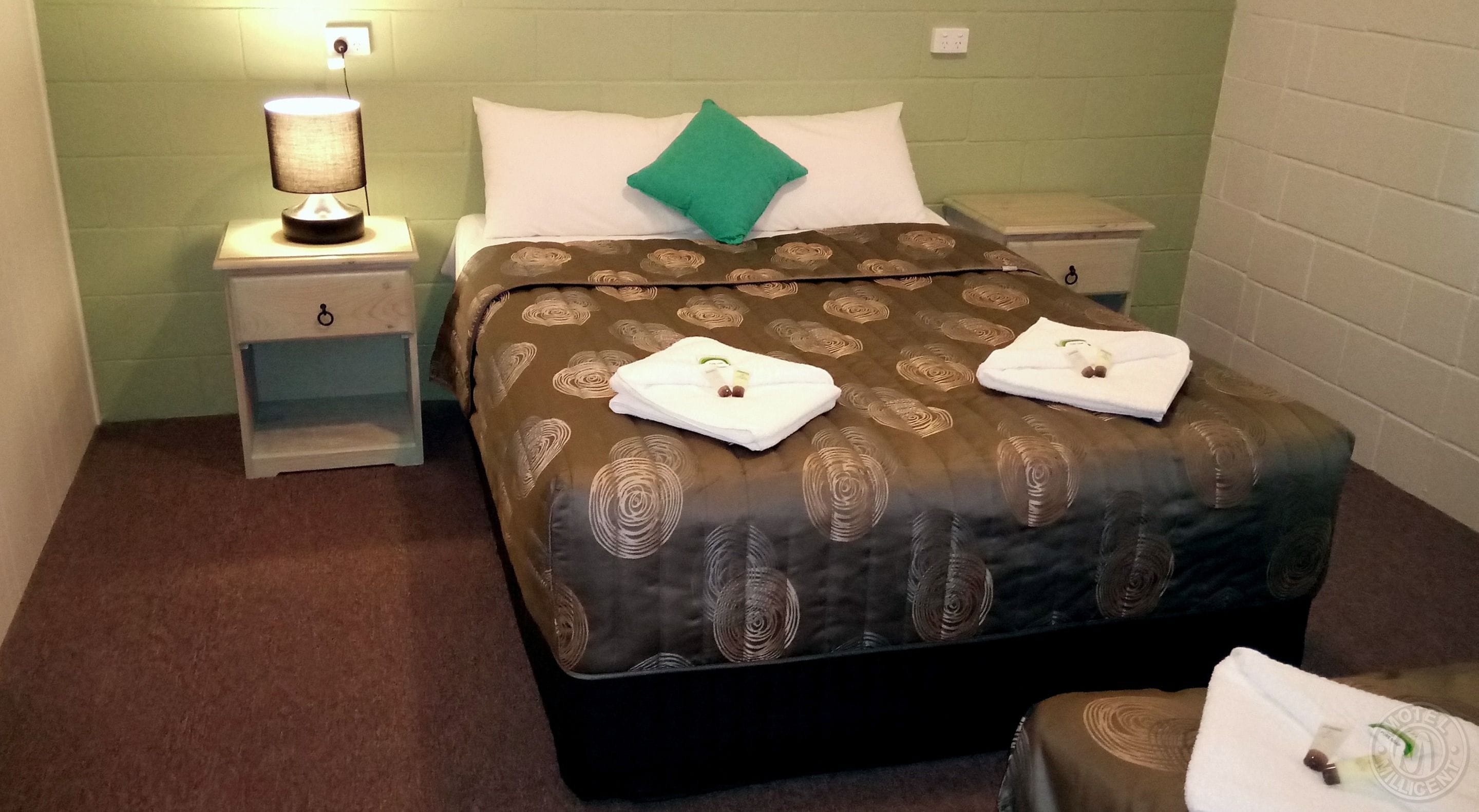 Cama bem arrumada com colcha estampada, um travesseiro verde e duas toalhas dobradas. Tem uma mesinha de cabeceira com uma luminária e uma gavetinha. O quarto tem paredes verdes e piso coberto por carpete.