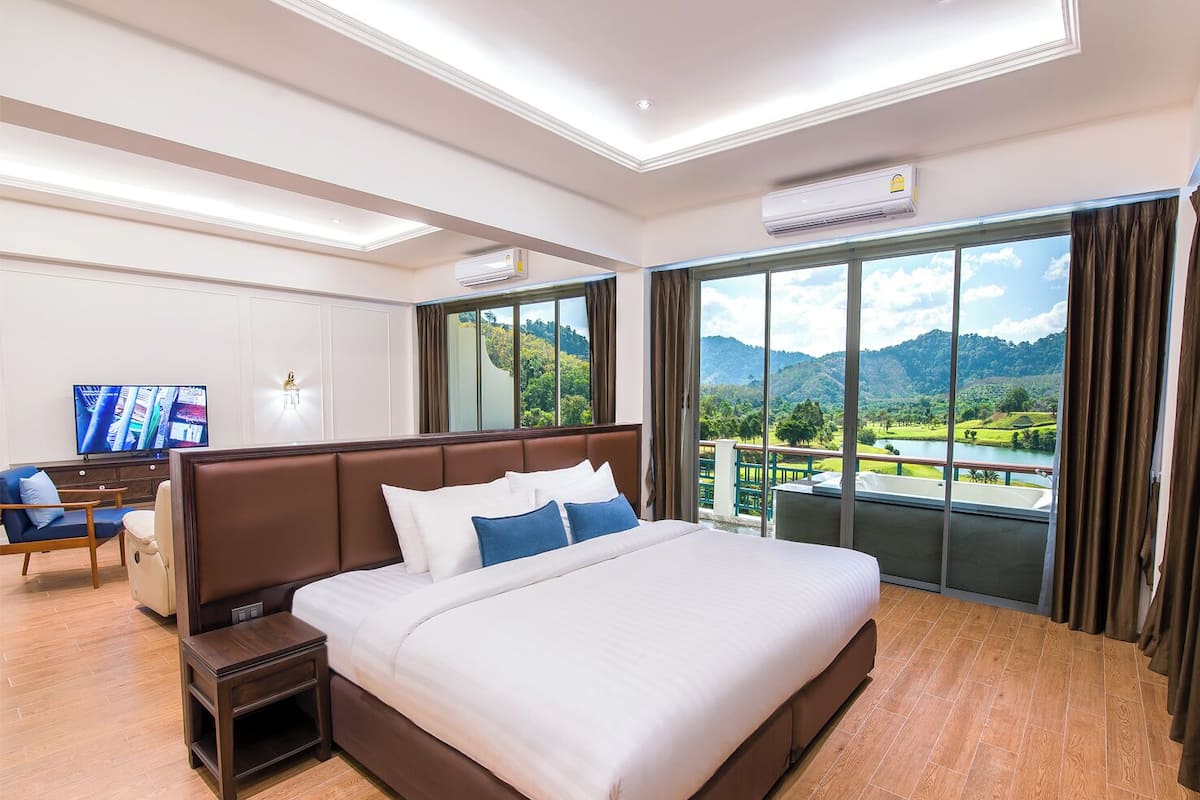 Quarto de hotel com uma cama grande, televisão, escrivaninha e vista para um lago e montanhas.