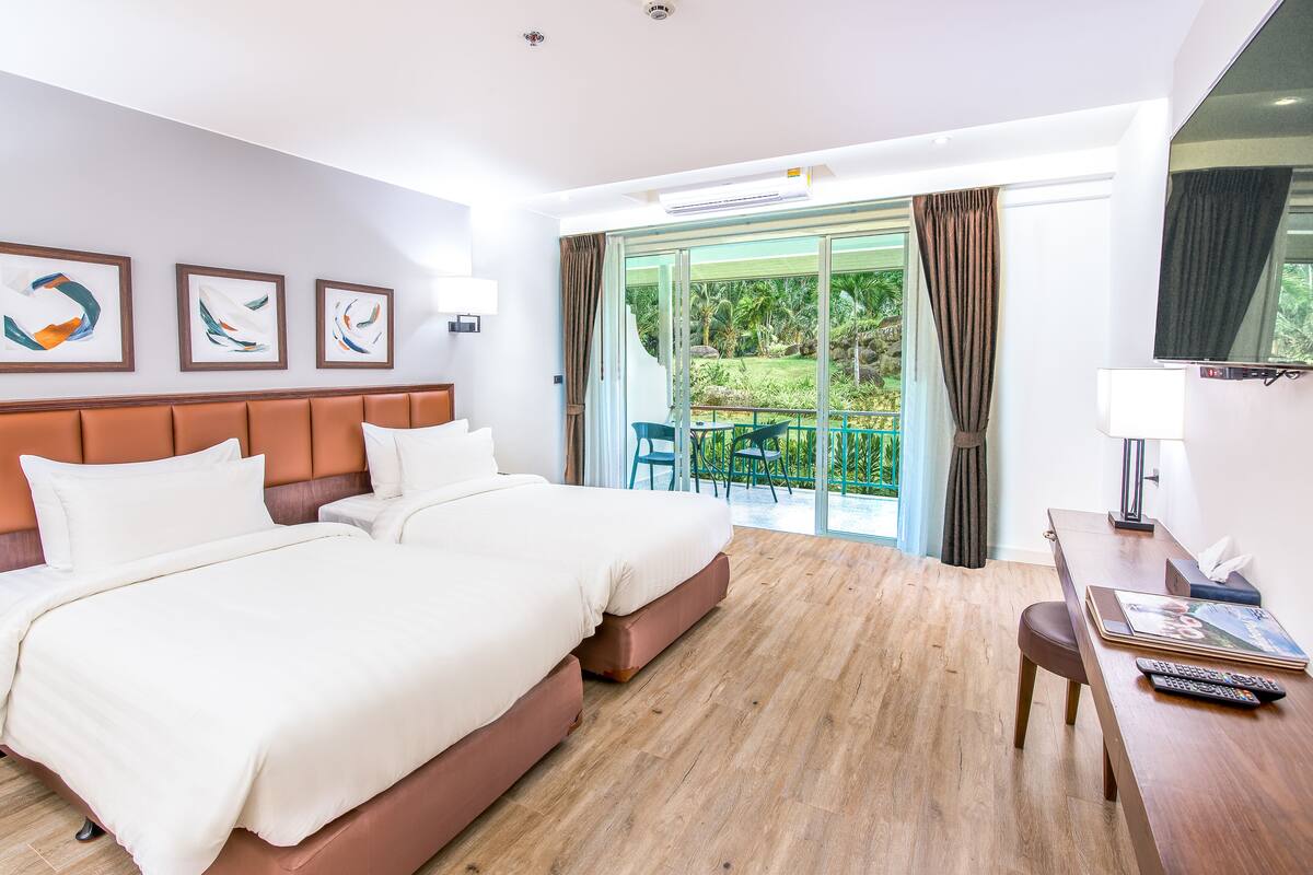 Quarto de hotel com uma cama grande, uma escrivaninha com cadeira, uma televisão e uma varanda com vista para o verde.