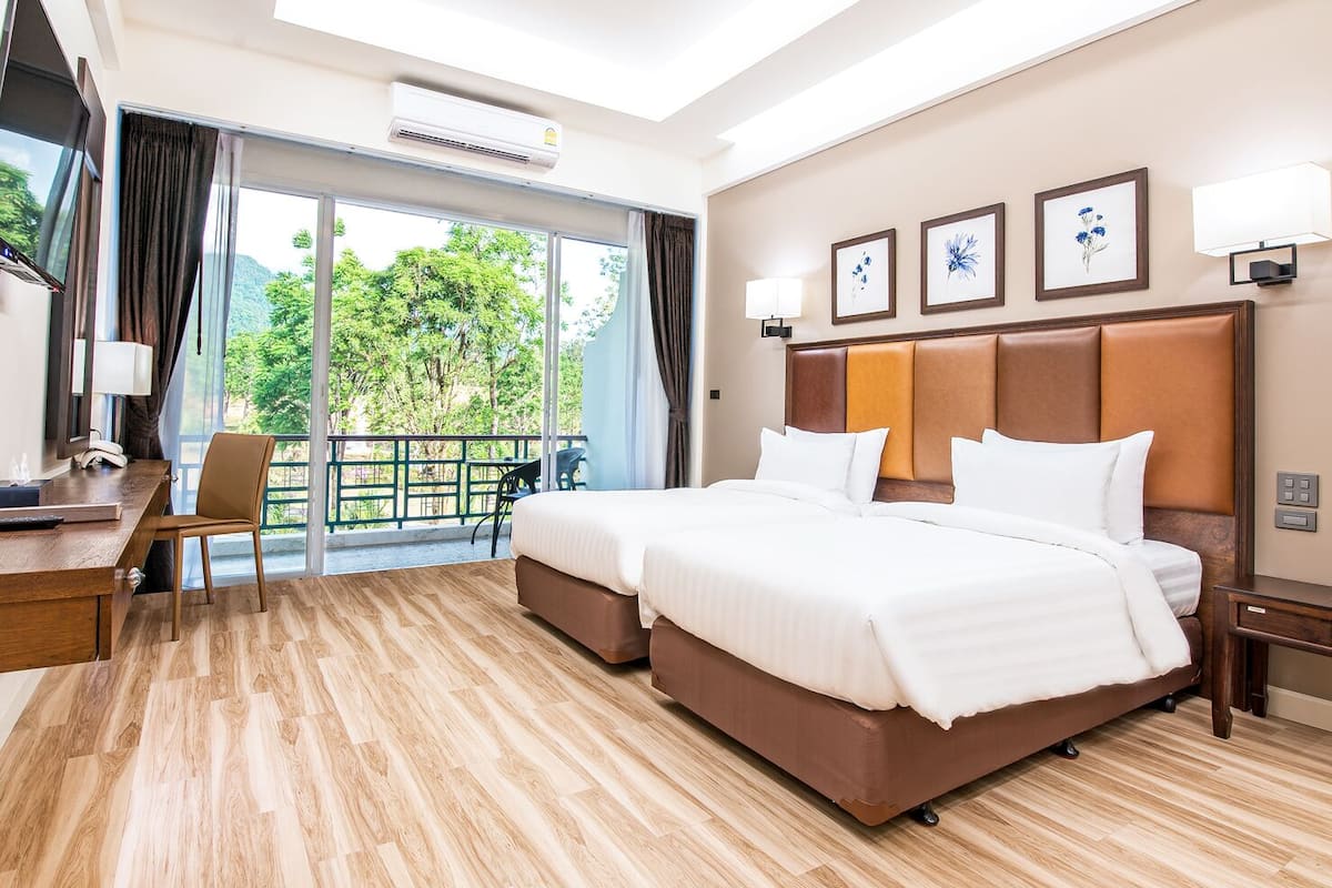 Quarto de hotel com uma cama grande, uma escrivaninha e uma cadeira. Possui uma varanda com vista para árvores e um edifício.