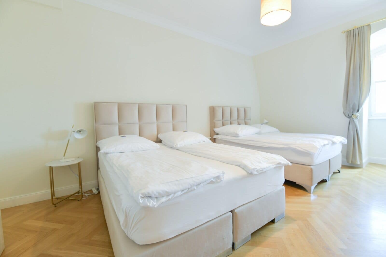 Quarto de hotel com duas camas, uma mesa de cabeceira, uma luminária e uma janela com cortinas.