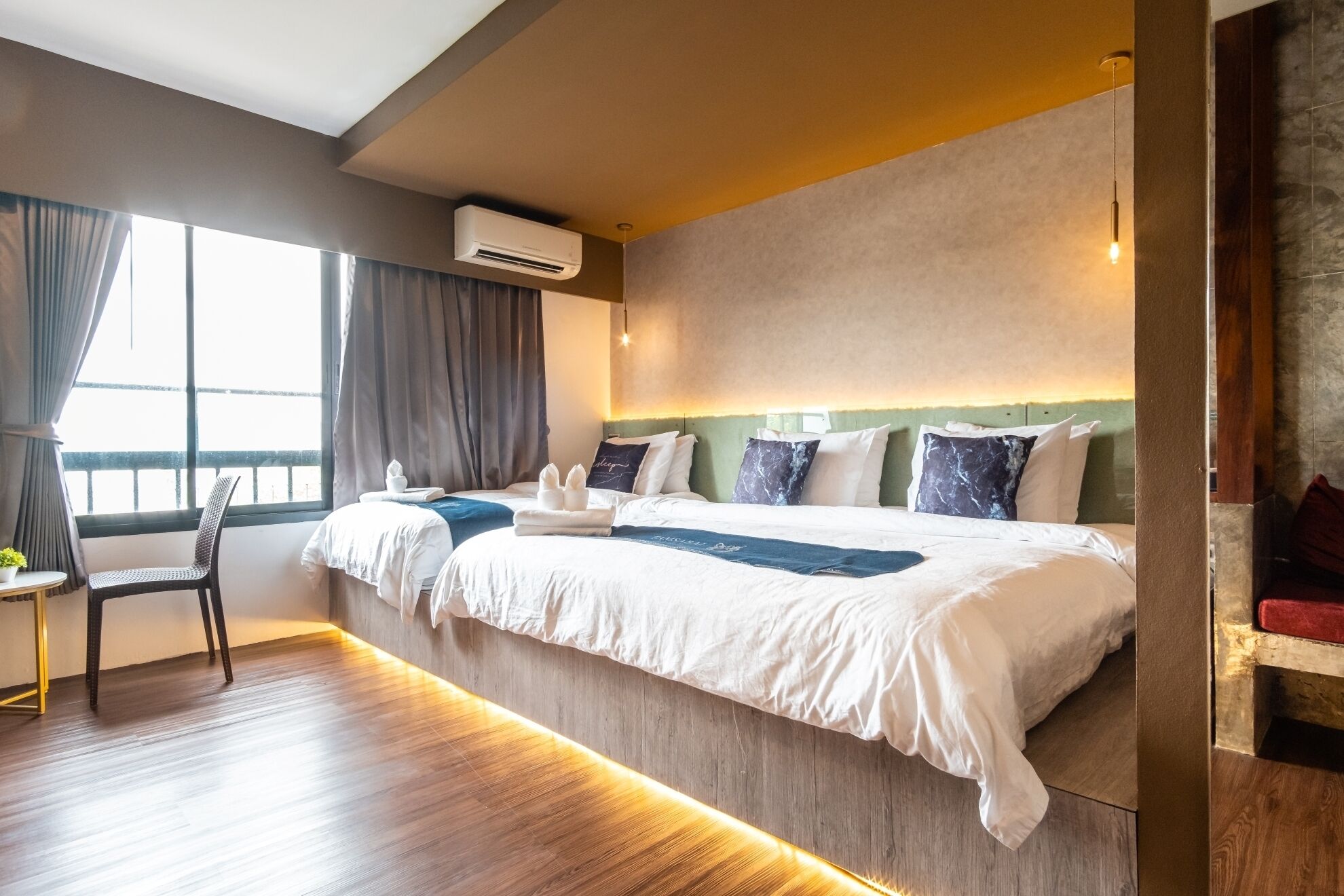 Quarto de hotel com duas camas, uma cadeira, ar condicionado e uma janela com cortinas.