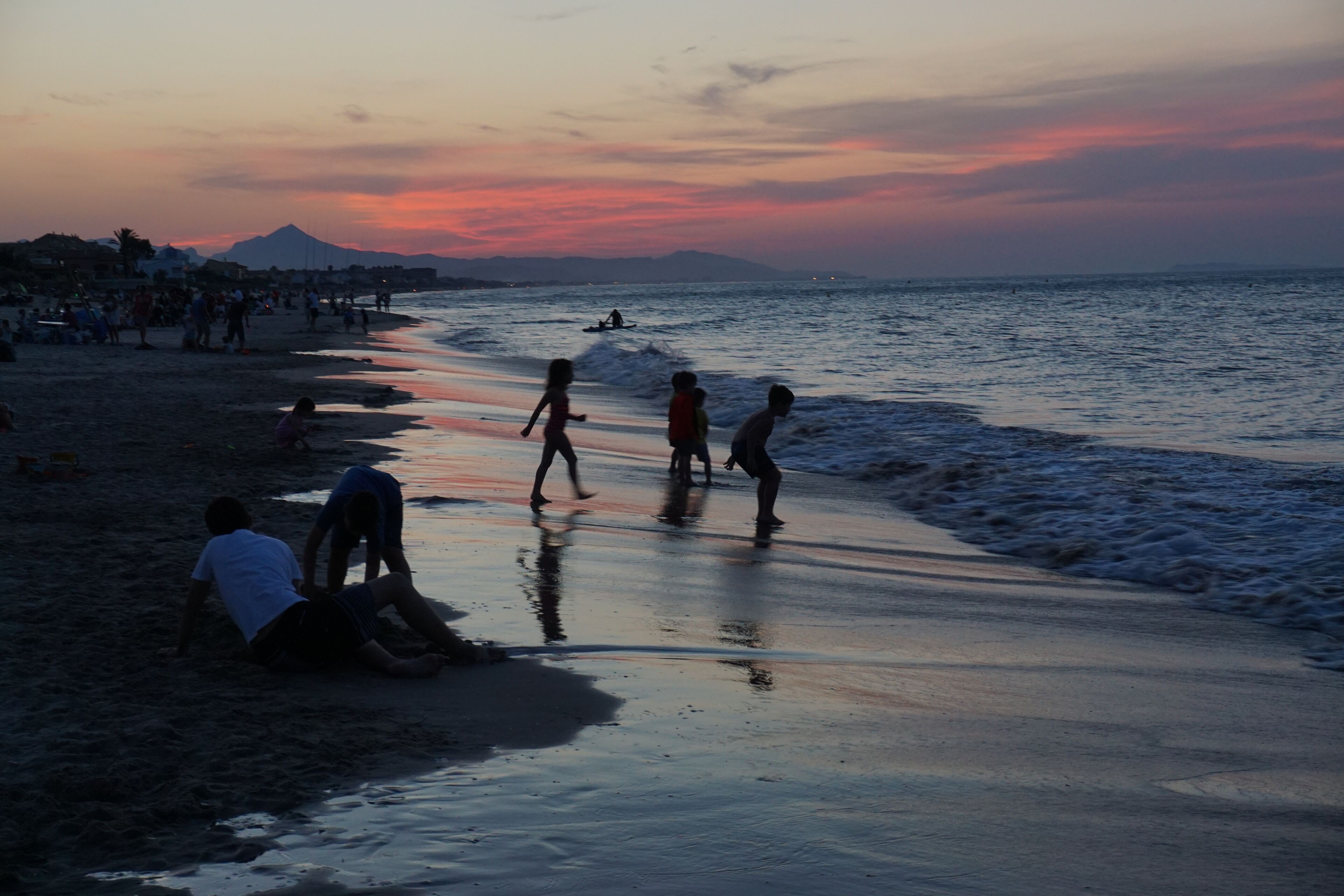 Cena de praia com pessoas caminhando pela orla, uma montanha ao fundo e um pôr do sol.