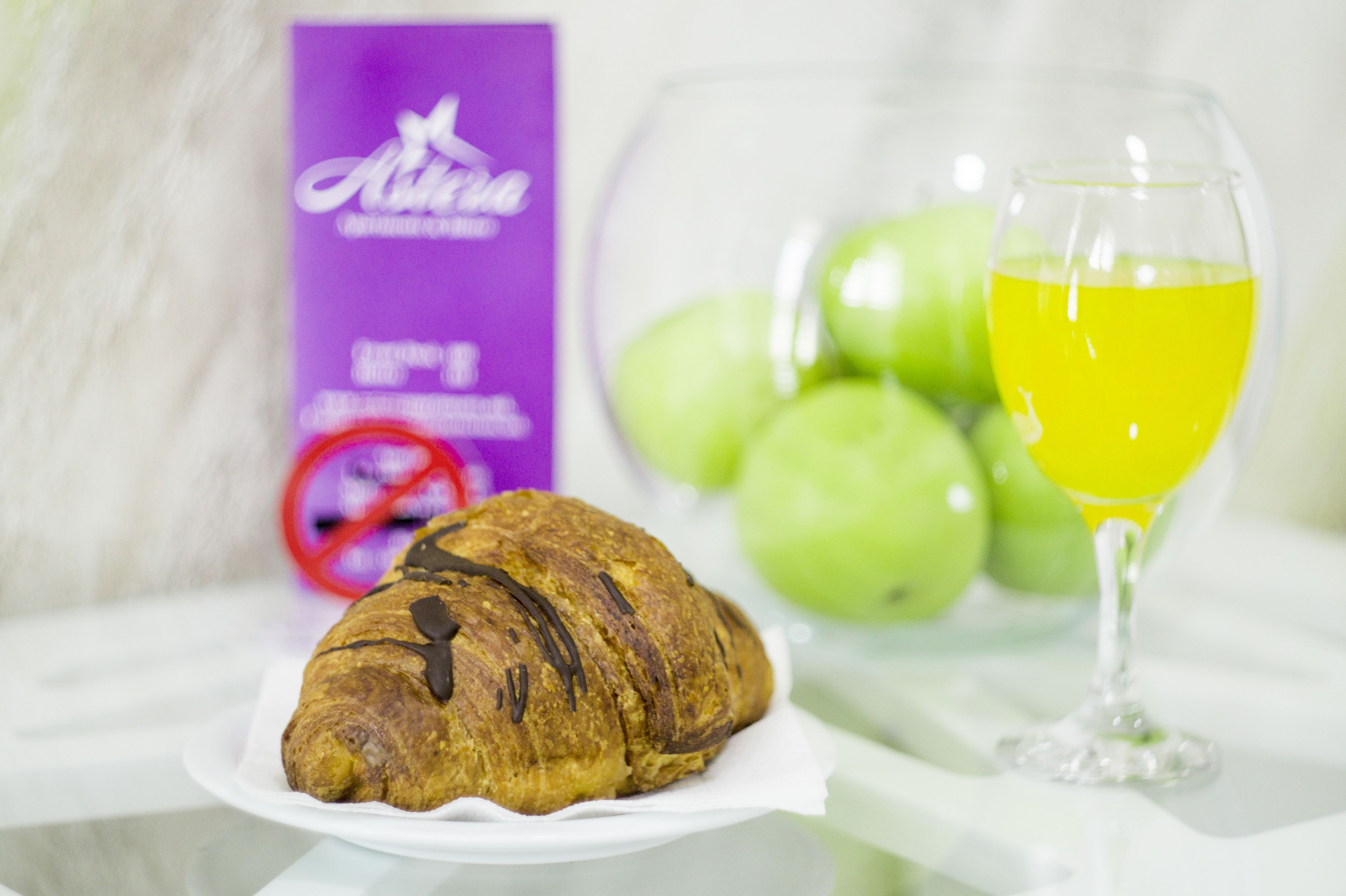 Um croissant sobre um prato branco, um copo de líquido amarelo e maçãs verdes ao fundo.